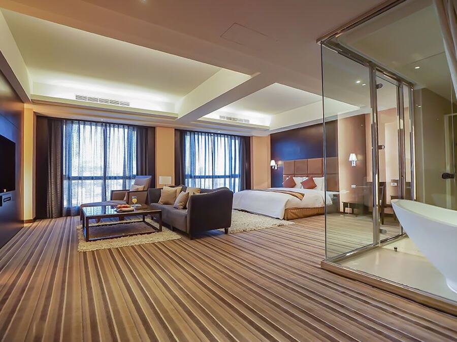 Quarto de hotel moderno com uma cama grande, um sofá, uma mesa de centro e um banheiro com paredes de vidro.