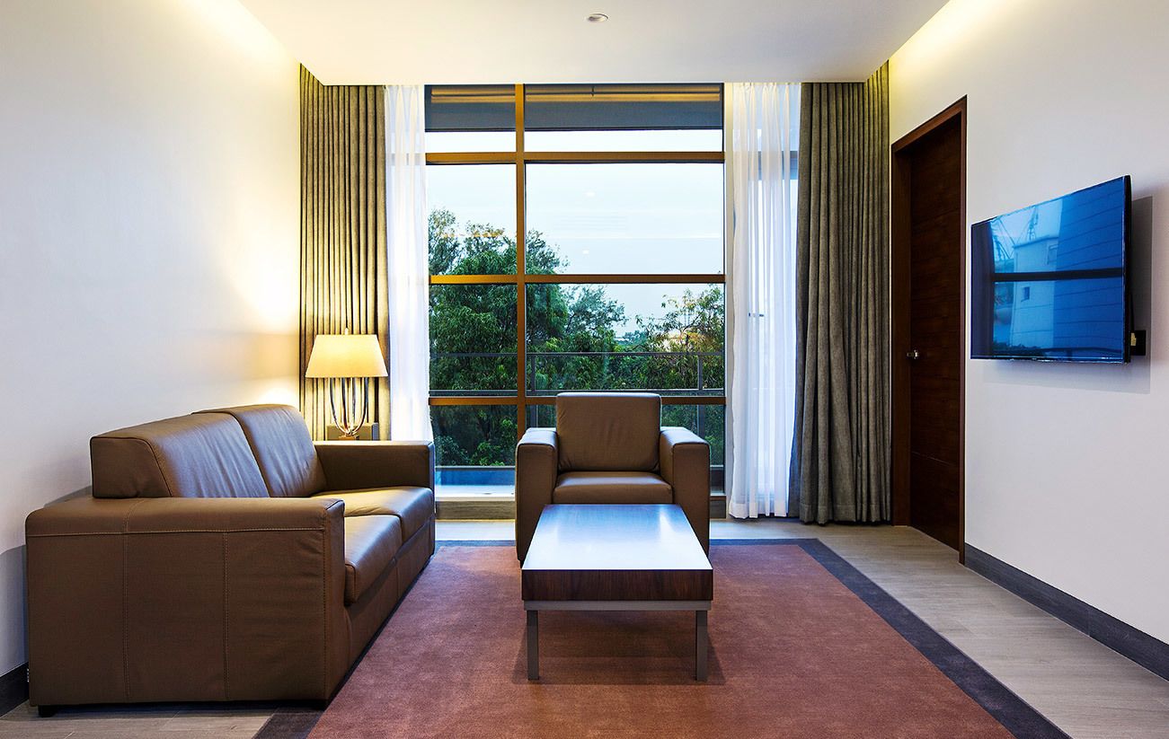 Quarto de hotel moderno com sofá, poltrona e mesa de centro. Uma televisão está fixada na parede e há uma janela grande com cortinas.