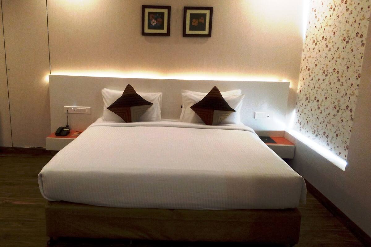 Quarto de hotel com uma cama grande, duas mesas de cabeceira, um criado-mudo com telefone e uma luminária na parede.