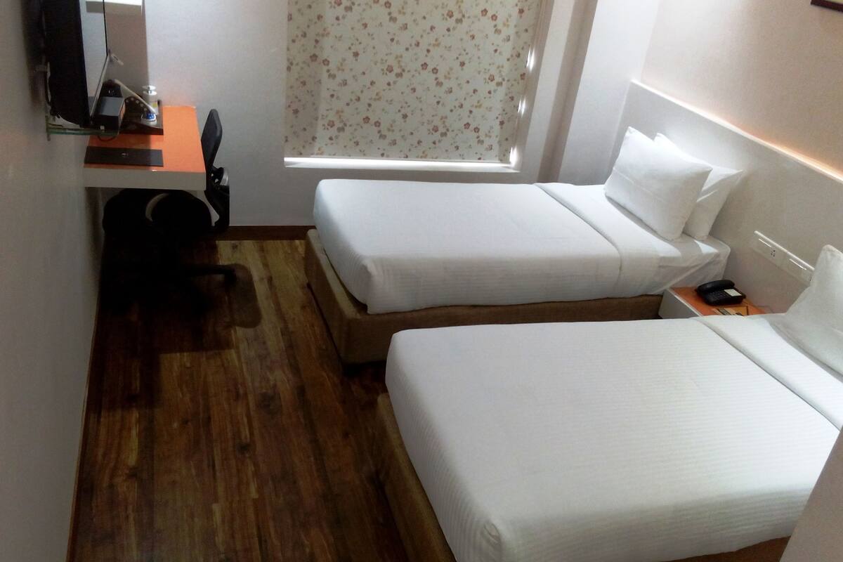 Quarto de hotel pequeno com duas camas de solteiro, uma escrivaninha, uma cadeira, televisão e janela com cortinas florais.