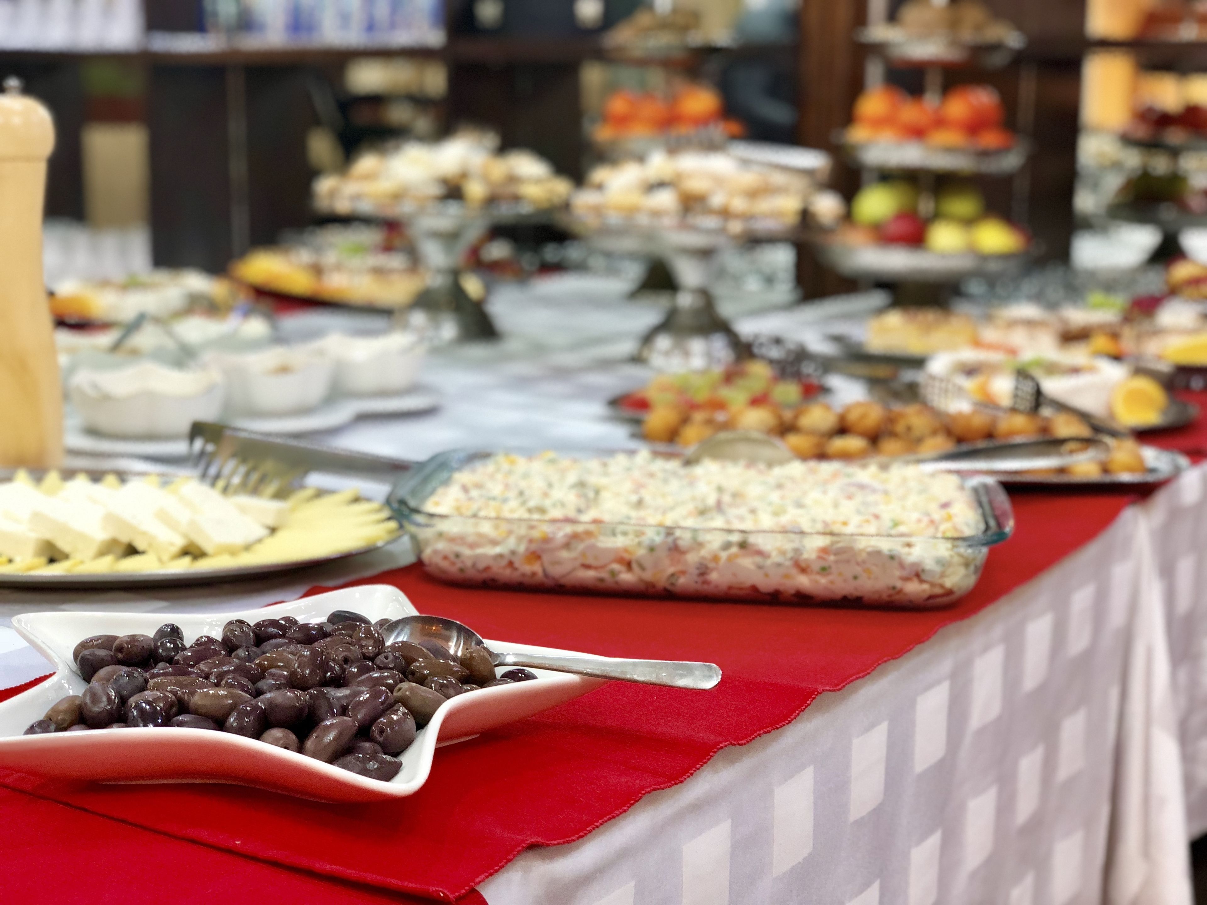 Mesa de buffet com diversos alimentos, incluindo um prato de azeitonas, um assado e várias travessas de frutas e legumes.