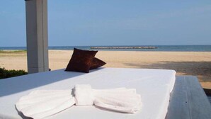 Cama com roupa de cama branca e toalha dobrada, travesseiro escuro, e vista para a praia e oceano.