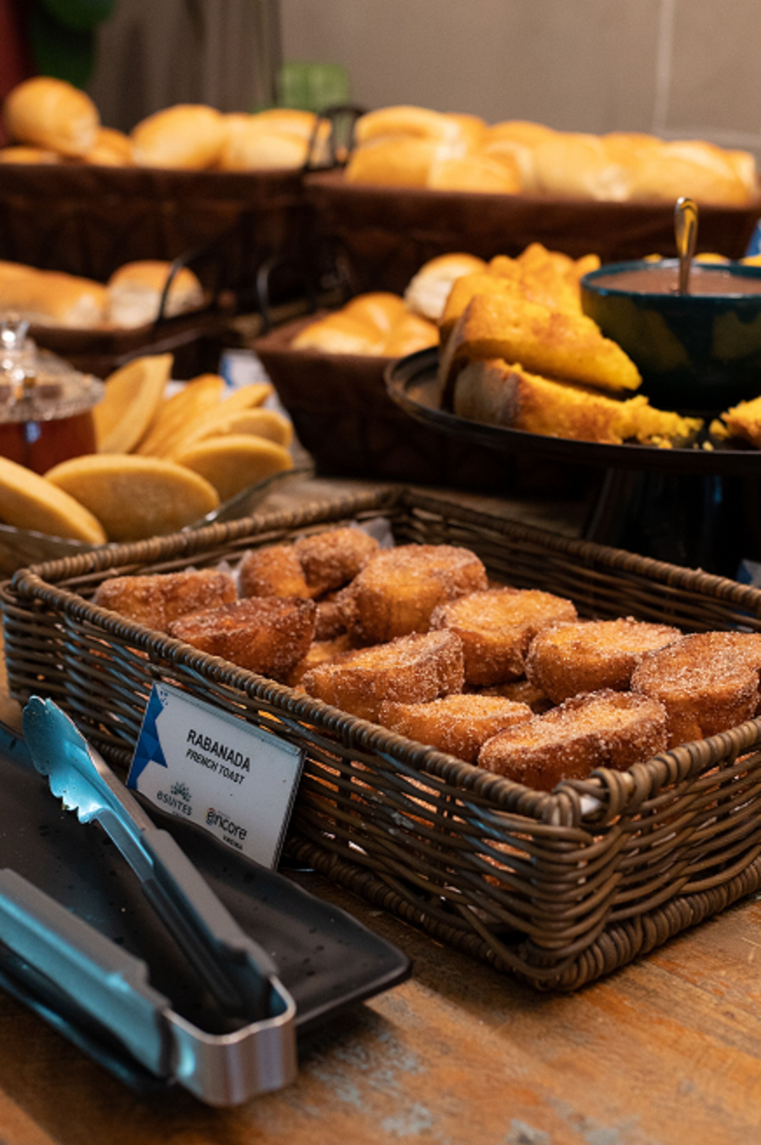 Buffet com diversos alimentos, incluindo pão, doces e uma placa indicando 'RABANADA FRANCE TOAST'.