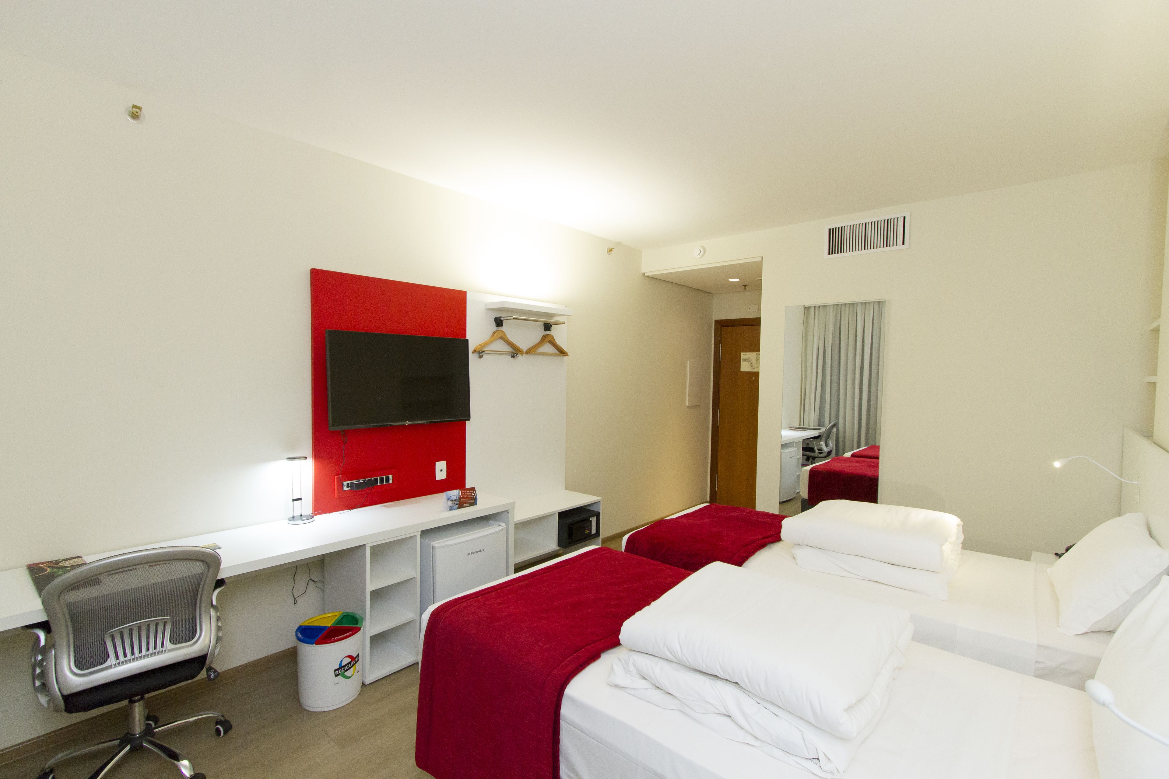 Quarto de hotel com duas camas, uma escrivaninha, uma cadeira, uma televisão e um guarda-roupa.