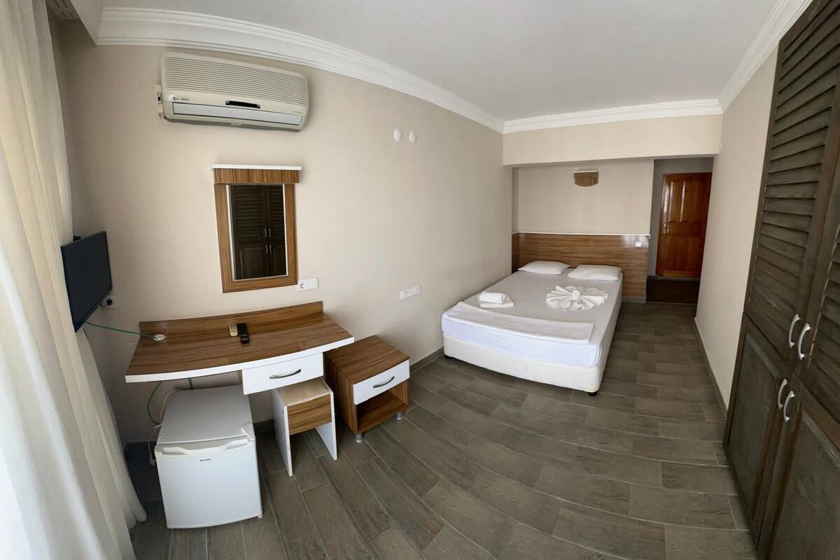 Quarto de hotel compacto com cama, escrivaninha, cadeira, pequeno frigorífico e televisão.