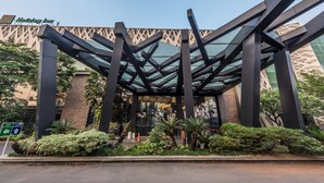 Exterior de um Holiday Inn, com entrada moderna em vidro e aço, rodeado por vegetação exuberante.