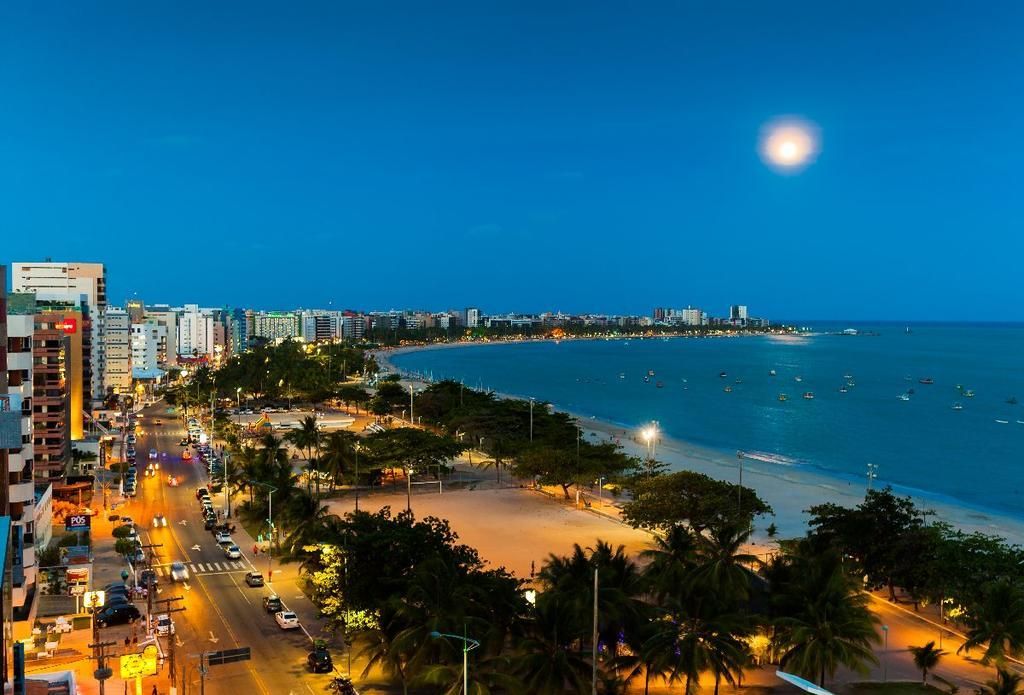 Paisagem urbana costeira à noite, com praia, edifícios e uma lua crescente.