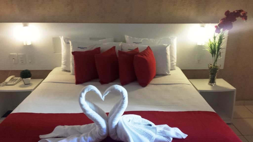 Quarto de hotel com cama, almofadas vermelhas, arranjo de toalha em formato de coração, um vaso com flores e uma mesa de cabeceira com telefone e uma pequena planta.