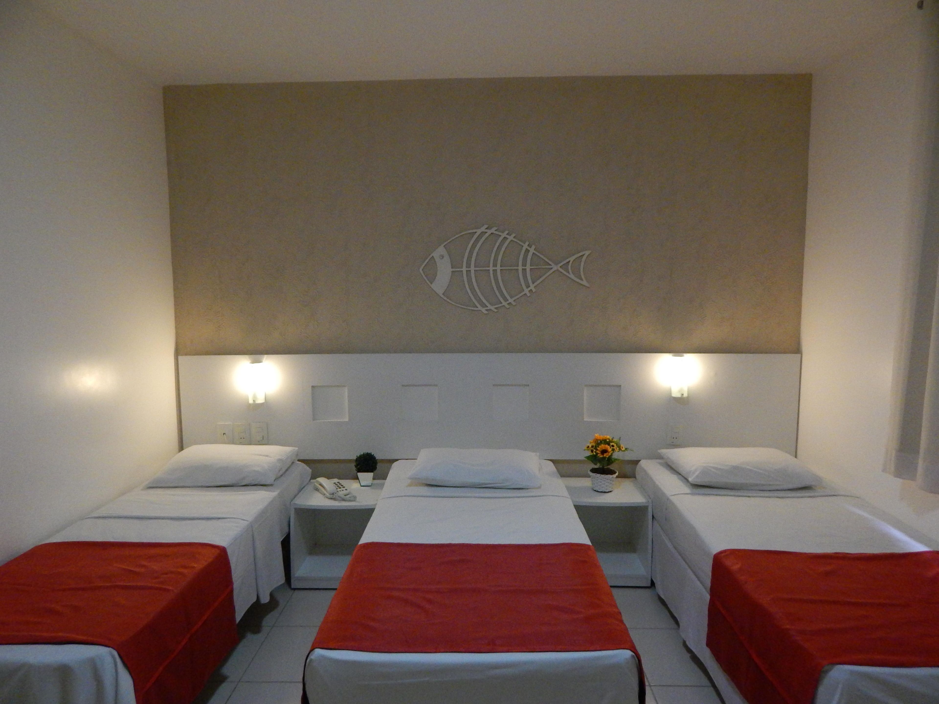 Quarto de hotel com três camas de solteiro, cada uma com colcha vermelha, cabeceira branca e decoração de peixe fixada na parede.
