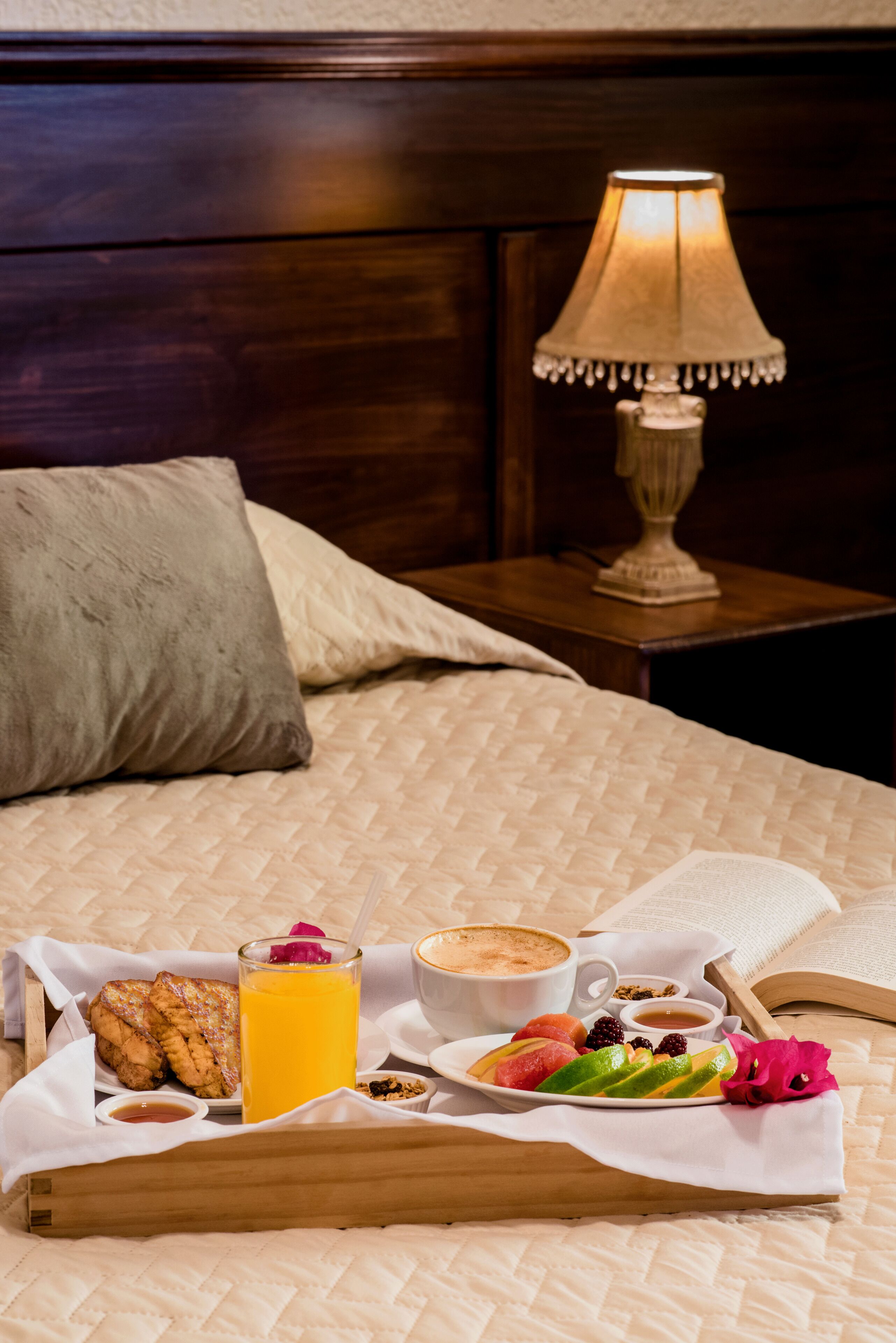 Cama bem arrumada com cabeceira de madeira, mesa de cabeceira com luminária e bandeja com itens de café da manhã, incluindo torradas, suco, café e frutas.