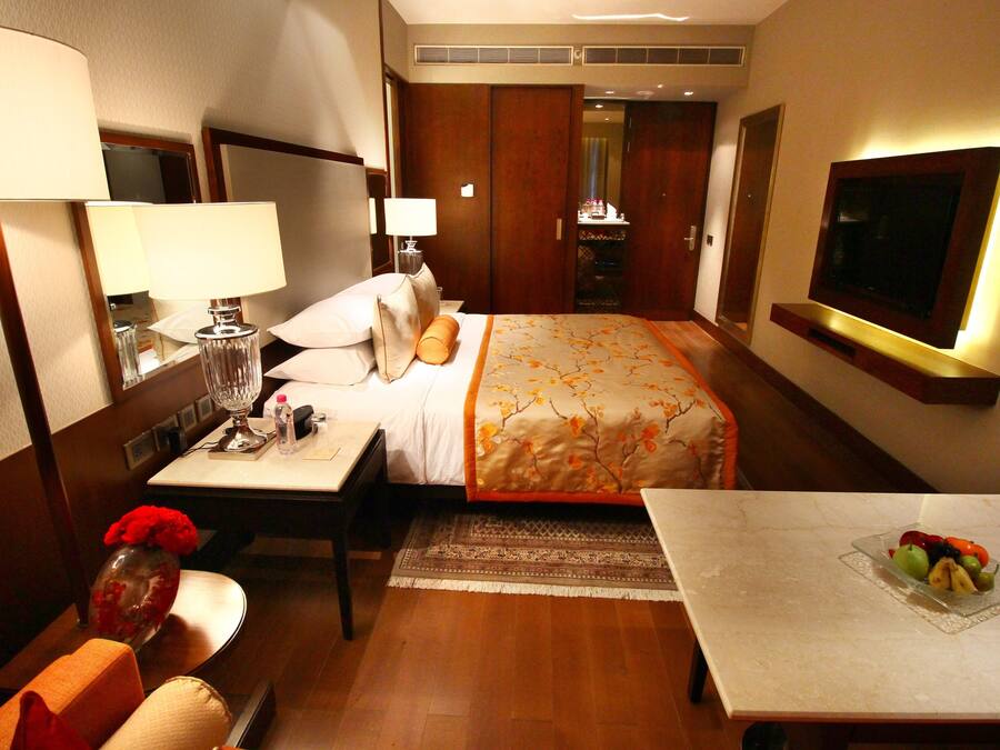 Quarto de hotel com cama, mesa de cabeceira, luminária, televisão e uma pequena mesa com frutas.