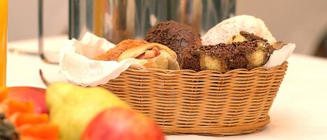 Uma mesa de café da manhã com cesta de palha cheia de doces, um prato de frutas e um bule de chá.