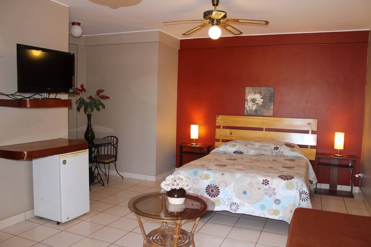 Quarto de hotel com cama, mesas de cabeceira, televisão, mini-geladeira, área de refeições e ventilador de teto.