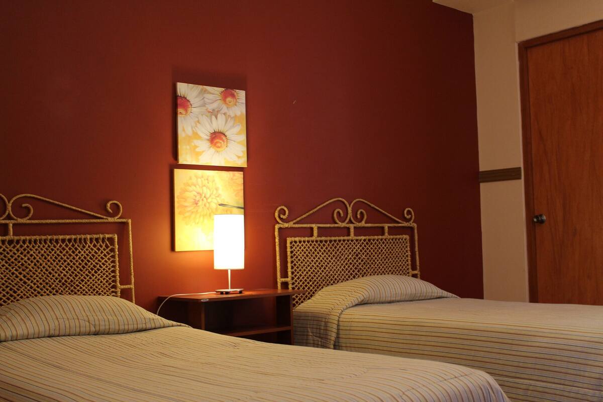 Quarto de hotel com duas camas, uma mesinha de cabeceira com abajur e uma parede com arte floral.