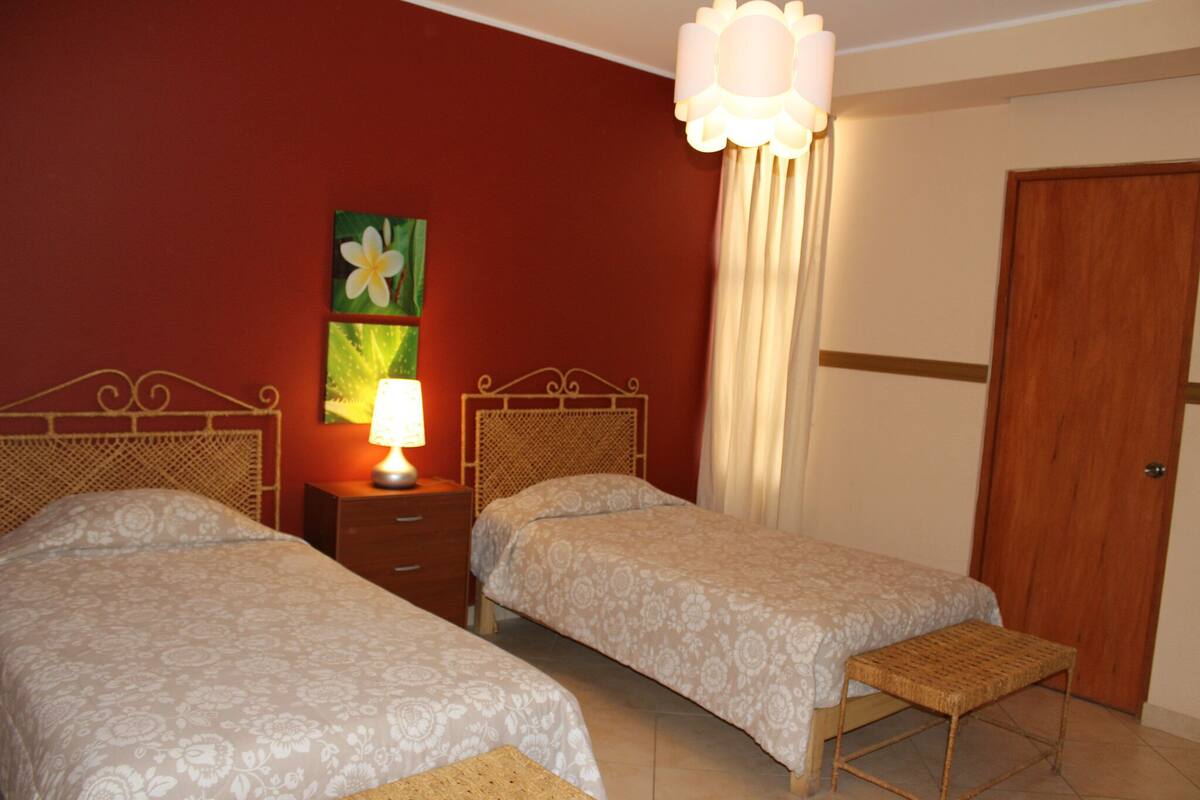 Quarto com duas camas, uma mesinha de cabeceira com luminária, uma porta de madeira e um quadro floral na parede.