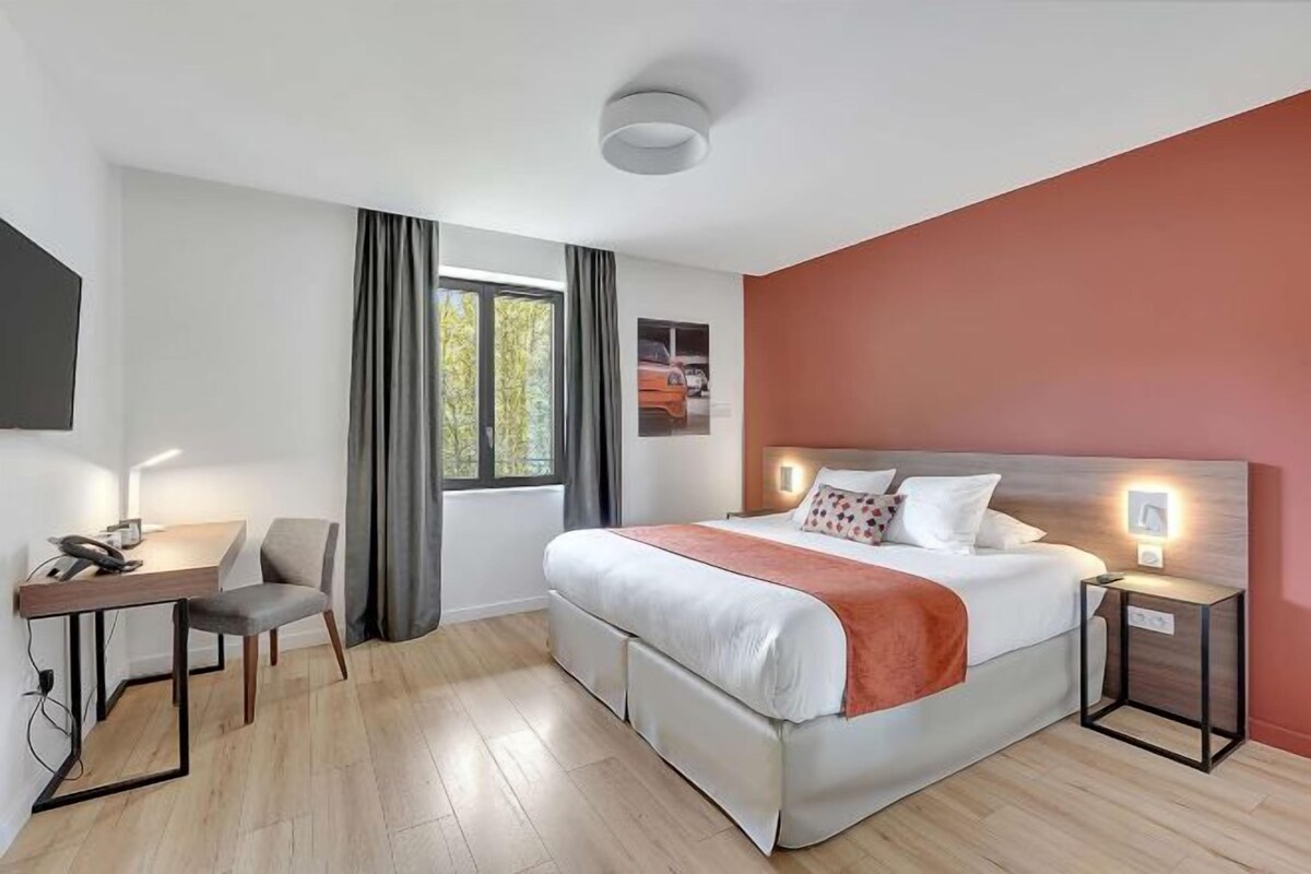 Quarto de hotel com cama, escrivaninha, cadeira e janela com cortinas.