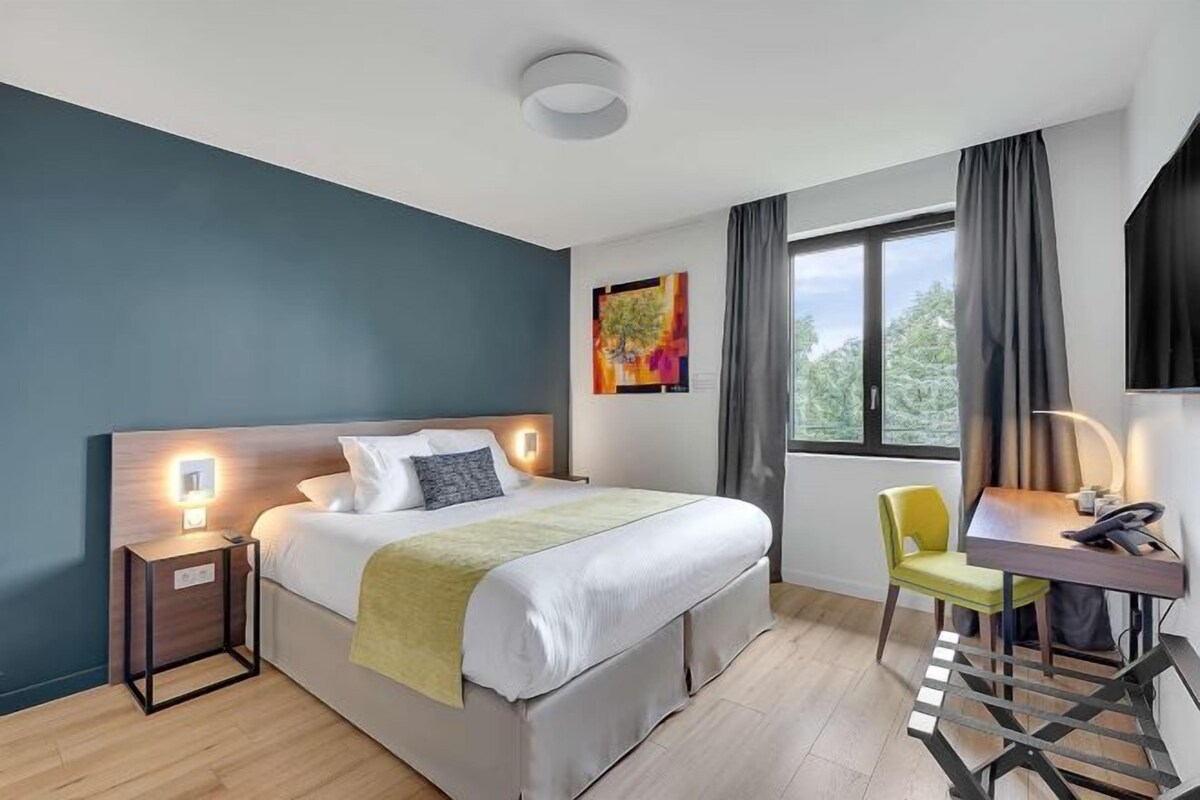 Quarto de hotel com cama, mesa de cabeceira, cadeira, escrivaninha e janela com cortinas.
