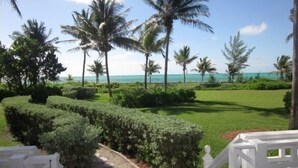Um jardim bem cuidado com um caminho que leva a um portão branco, rodeado por vegetação exuberante e palmeiras, com vista para um céu azul límpido e uma paisagem distante do oceano.