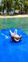Uma pessoa deitada em uma plataforma azul flutuante em água cristalina, cercada por palmeiras e uma praia de areia.