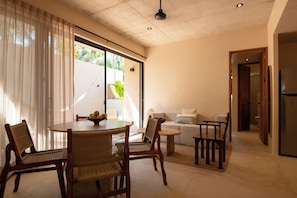 Uma sala de jantar com uma mesa redonda, quatro cadeiras e uma pequena mesa lateral. Há um sofá e uma janela com cortinas.