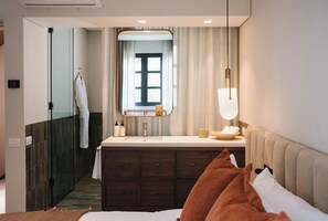Quarto de hotel com um espelho grande, toucador com lavatório, cama com almofadas castanhas e janela com cortinas.