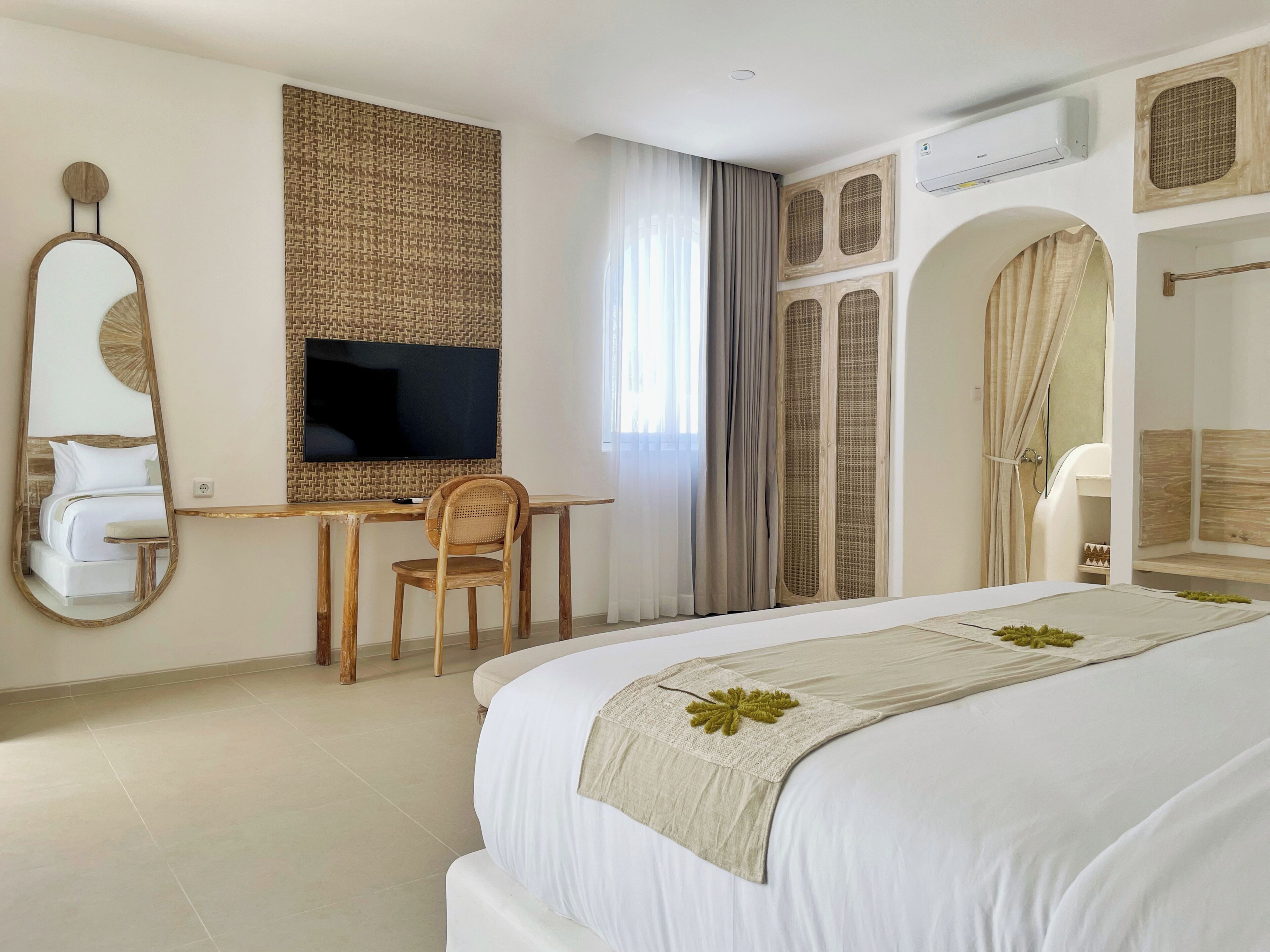 Quarto de hotel com uma cama, uma escrivaninha com cadeira, televisão e um espelho grande.