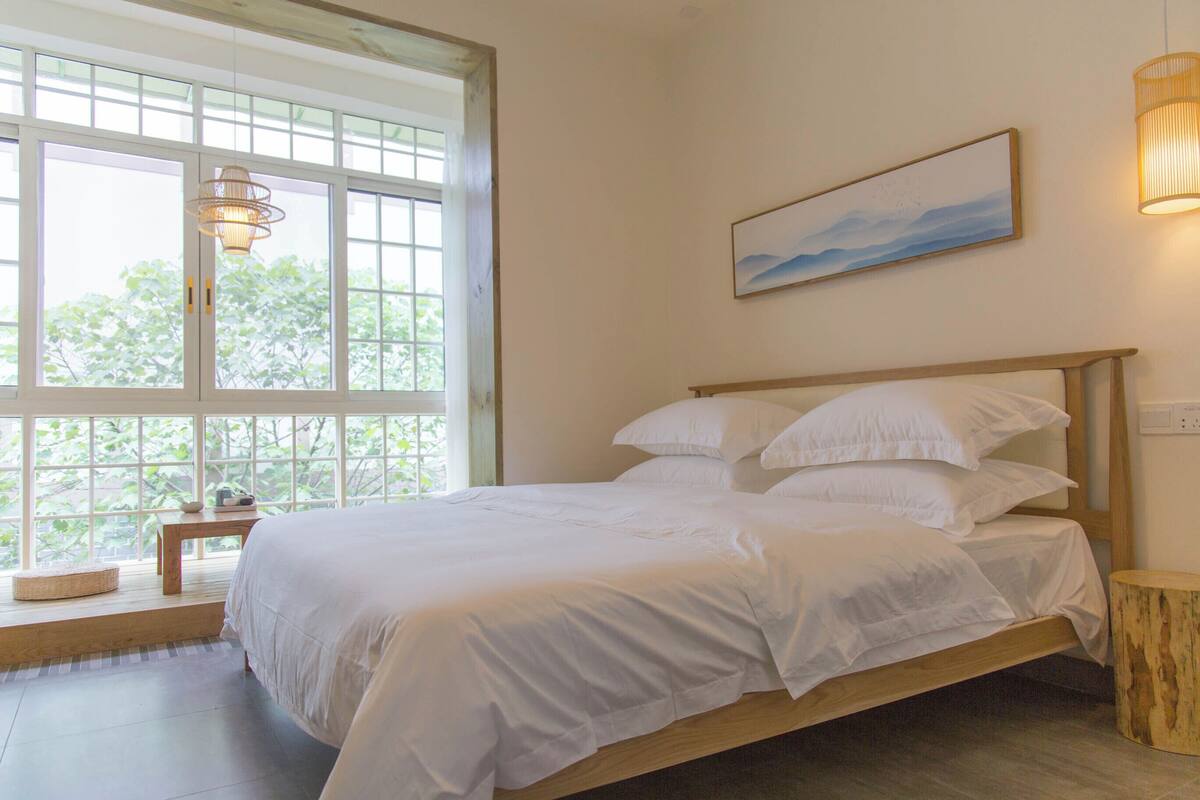 Quarto com uma janela grande, uma cama com roupa de cama branca, uma mesa de cabeceira e uma luminária fixada na parede.