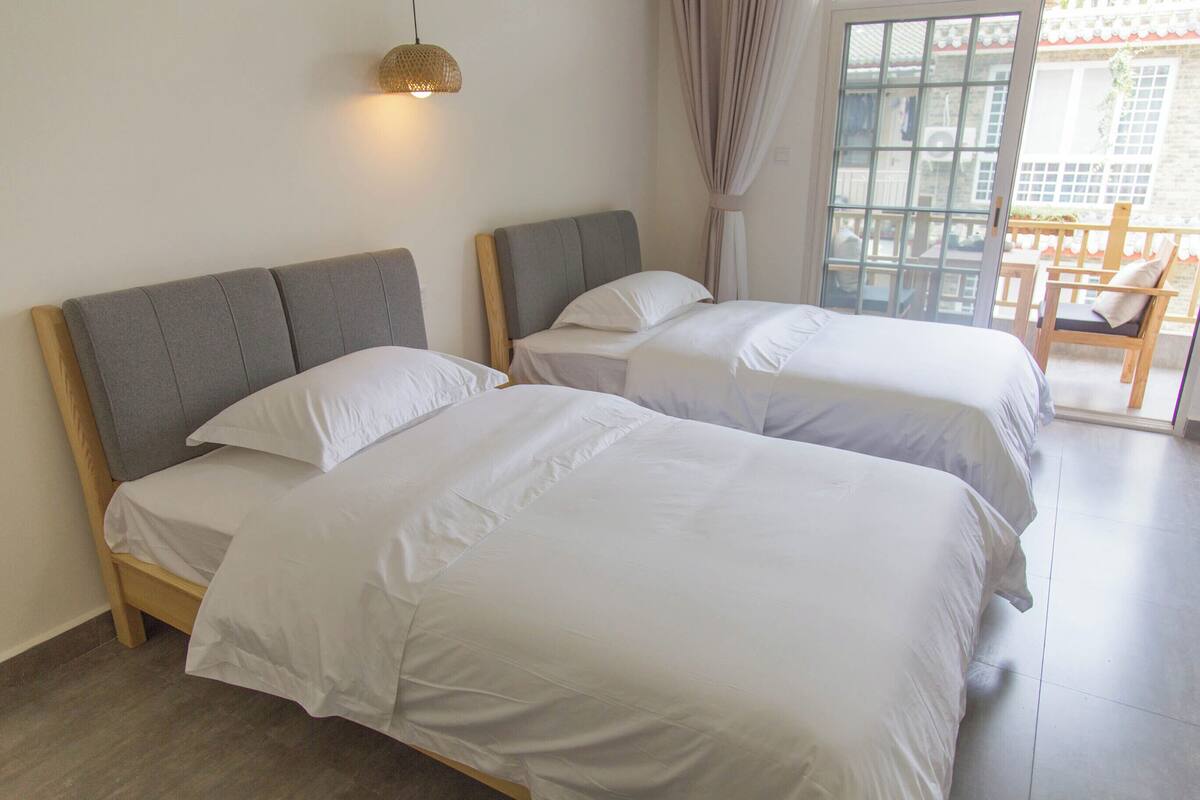 Quarto com duas camas, cabeceira de madeira, luminária trançada e vista por uma porta de vidro.