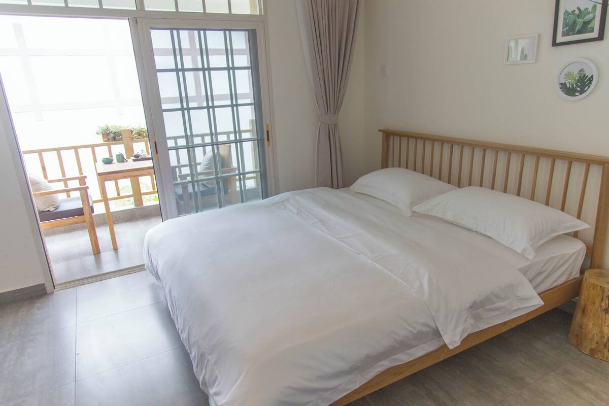 Quarto com cama de madeira, porta de vidro deslizante para uma varanda e um pequeno banquinho de madeira.