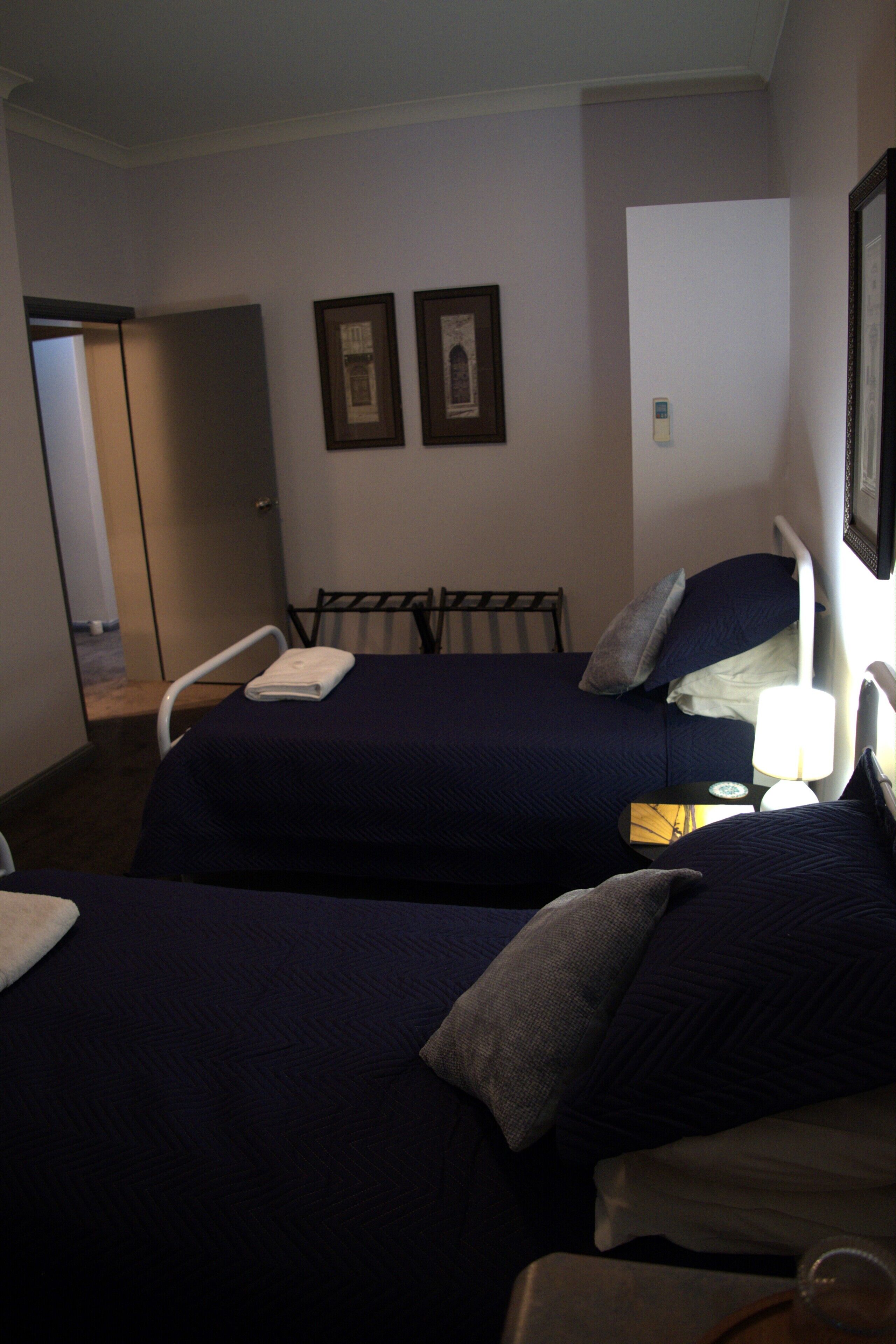 Quarto de hotel com duas camas, uma mesa de cabeceira com abajur e uma porta que dá para outro cômodo.