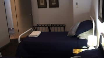 Quarto de hotel com duas camas, uma mesa de cabeceira com abajur e uma porta que dá para outro cômodo.