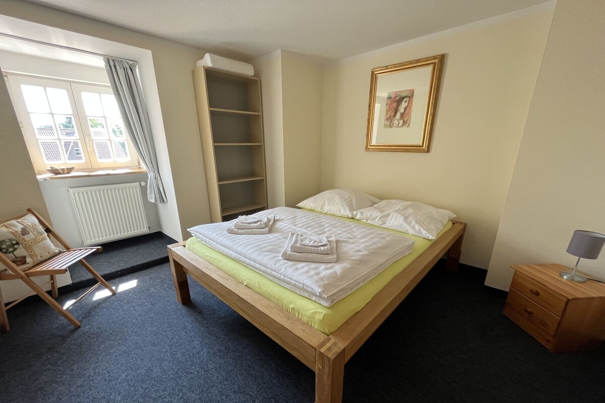 Um quarto com cama, mesa de cabeceira de madeira, cadeira, radiador, janela com cortinas e um quadro na parede.