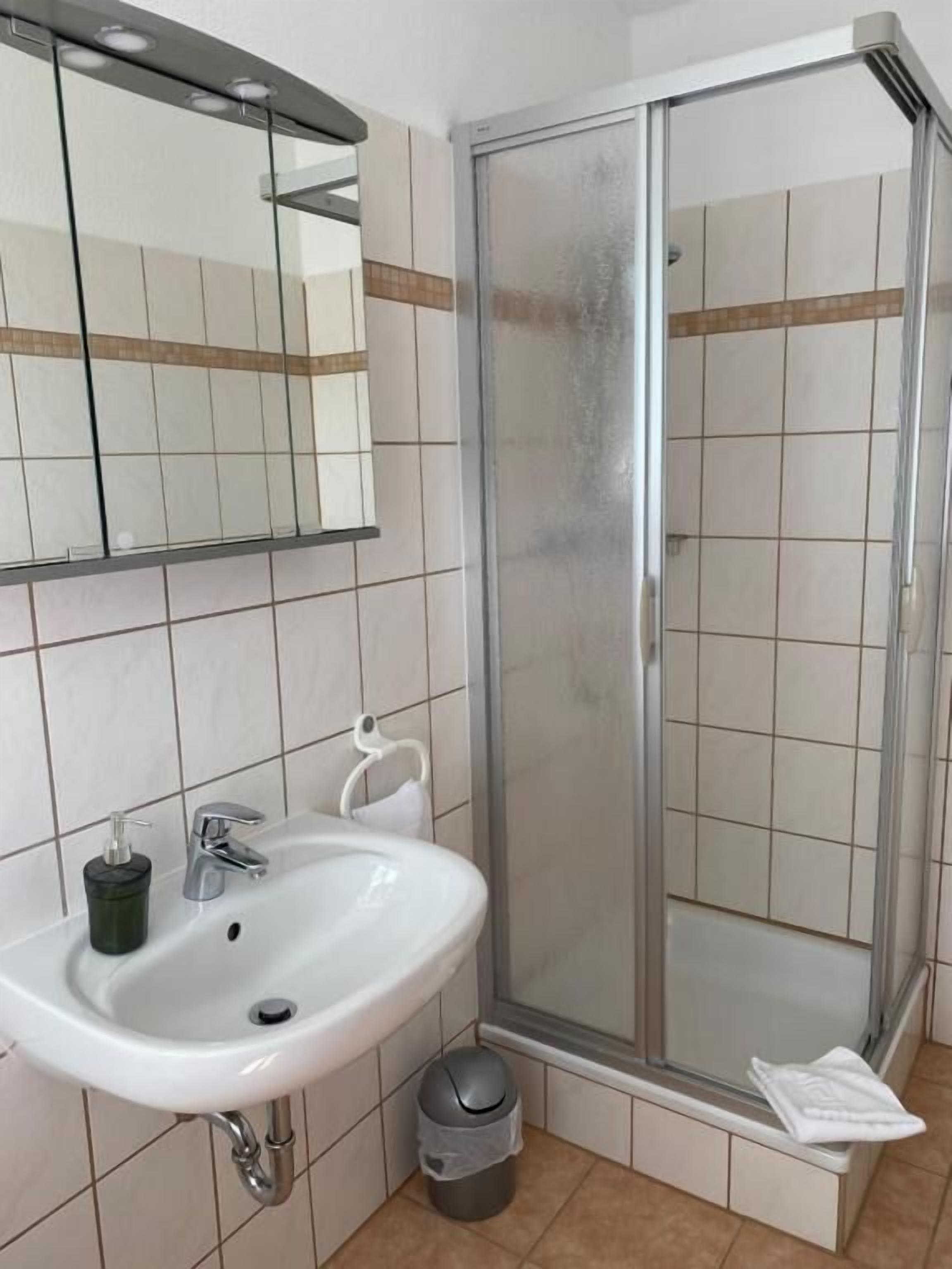 Casa de banho com lavatório branco, espelho, cabine de duche em vidro e um contentor de lixo.