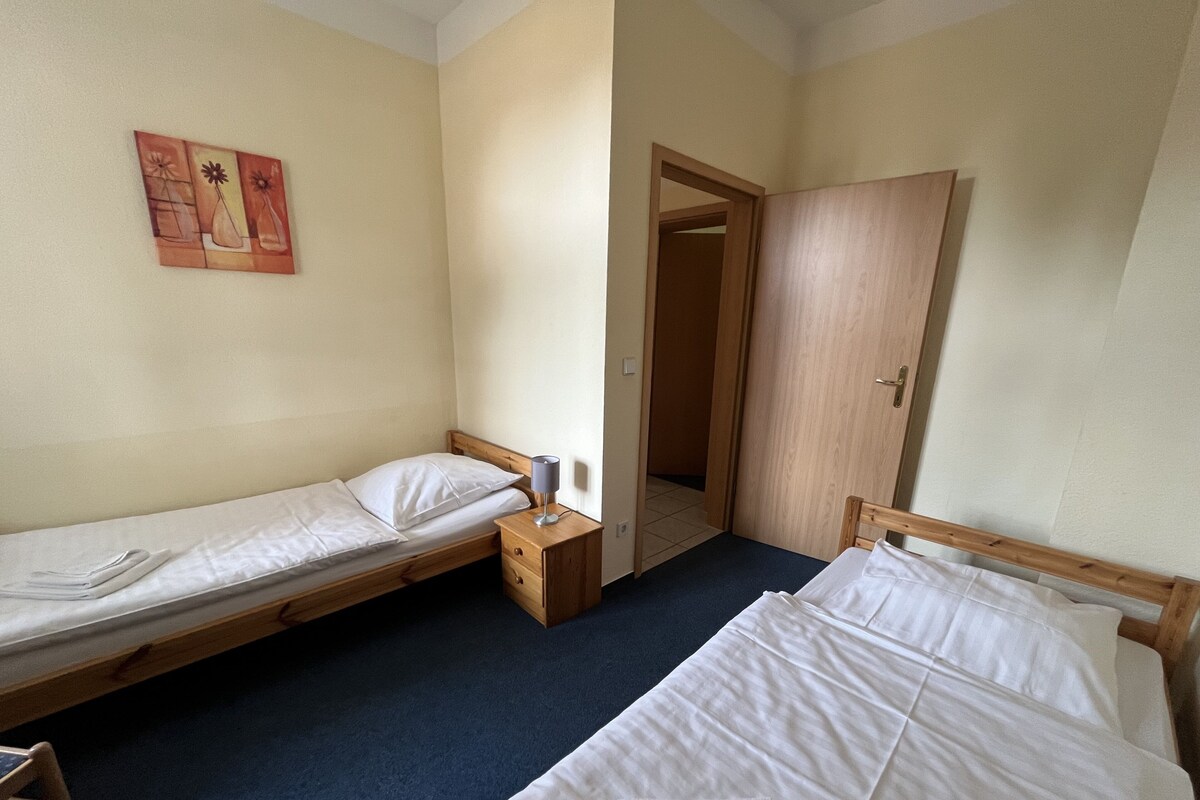 Quarto com duas camas de solteiro, uma mesa de cabeceira de madeira, uma luminária e uma porta que dá para outro cômodo.