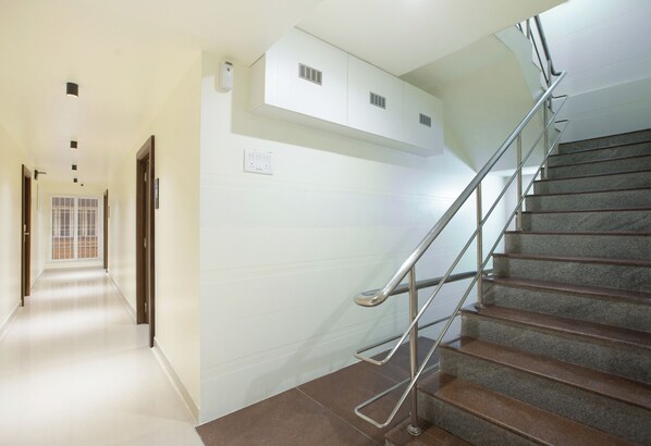 Uma escadaria com guarda-corpos de metal, um patamar com interruptor de luz e um corredor com uma porta e uma janela.