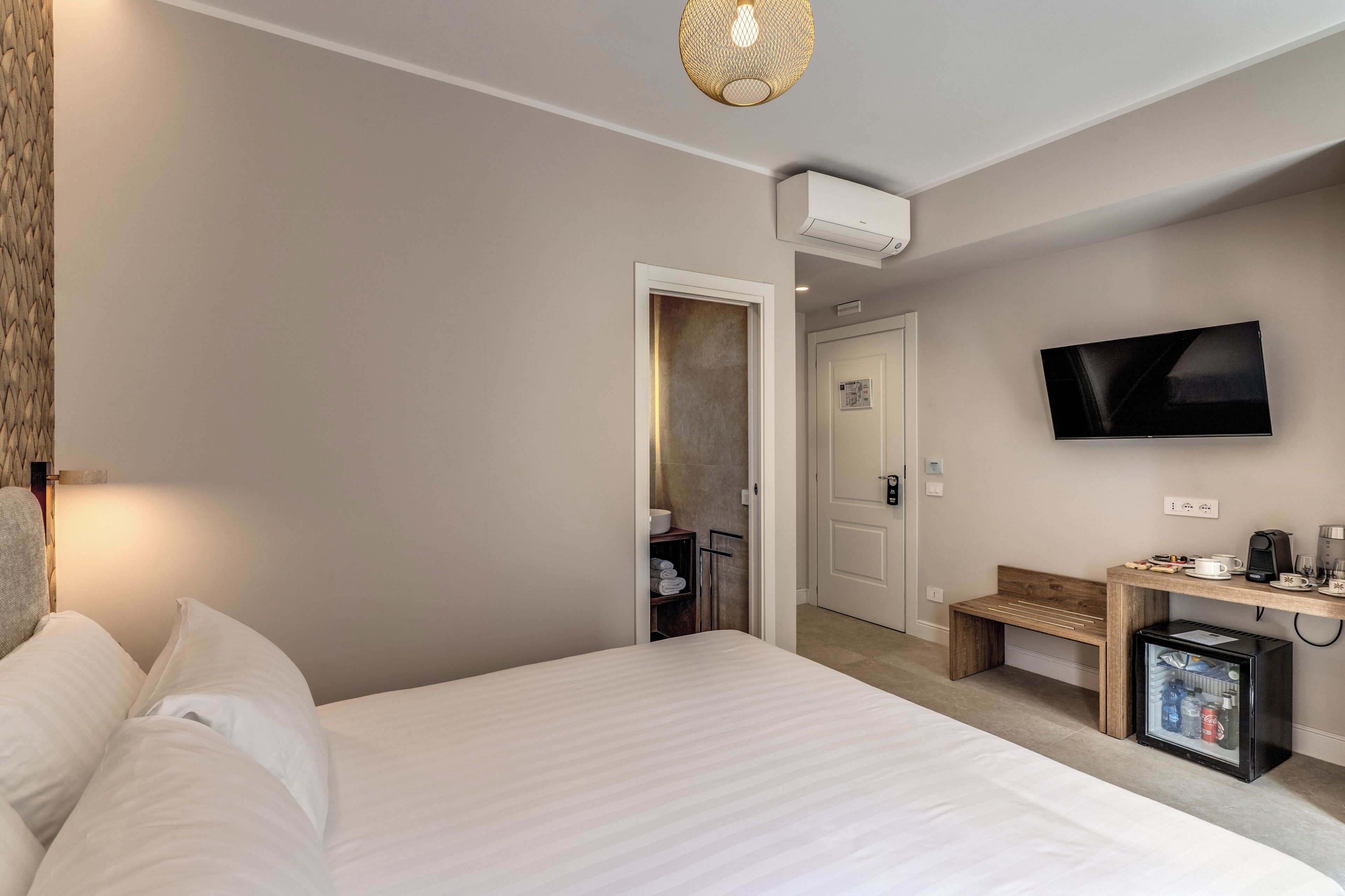 Quarto de hotel com uma cama, televisão, pequeno frigorífico, um banco e porta para uma casa de banho.