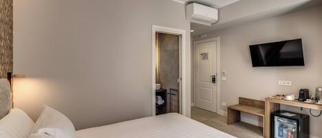 Quarto de hotel com uma cama, televisão, pequeno frigorífico, um banco e porta para uma casa de banho.
