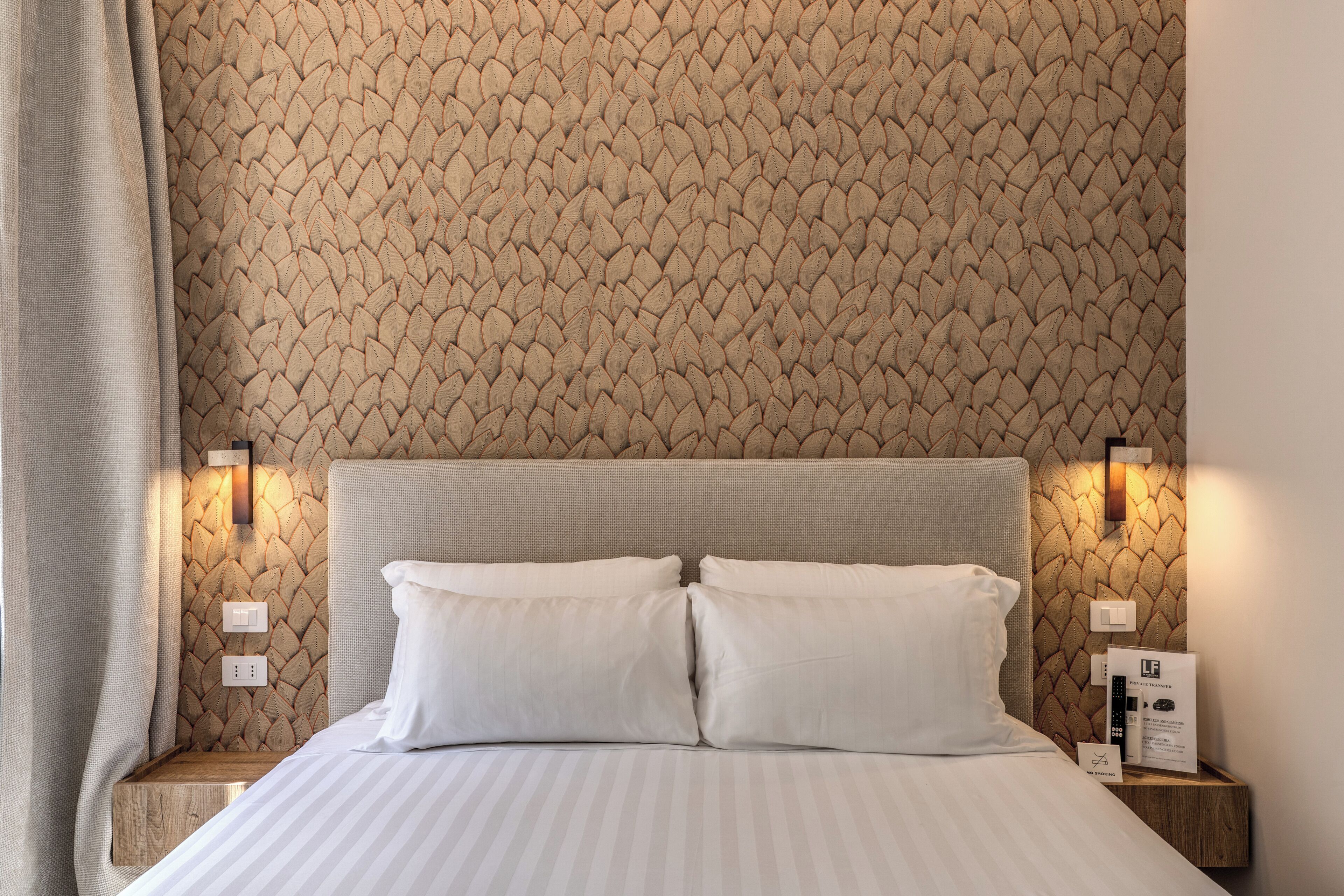 Quarto de hotel com parede estampada, duas luminárias de cabeceira, cama com roupa de cama branca e mesa de cabeceira com controle remoto.