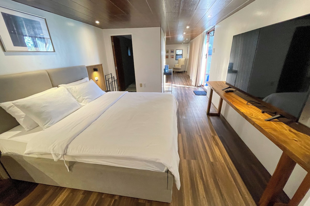 Quarto de hotel com uma cama grande, uma escrivaninha de madeira e vista para o corredor.