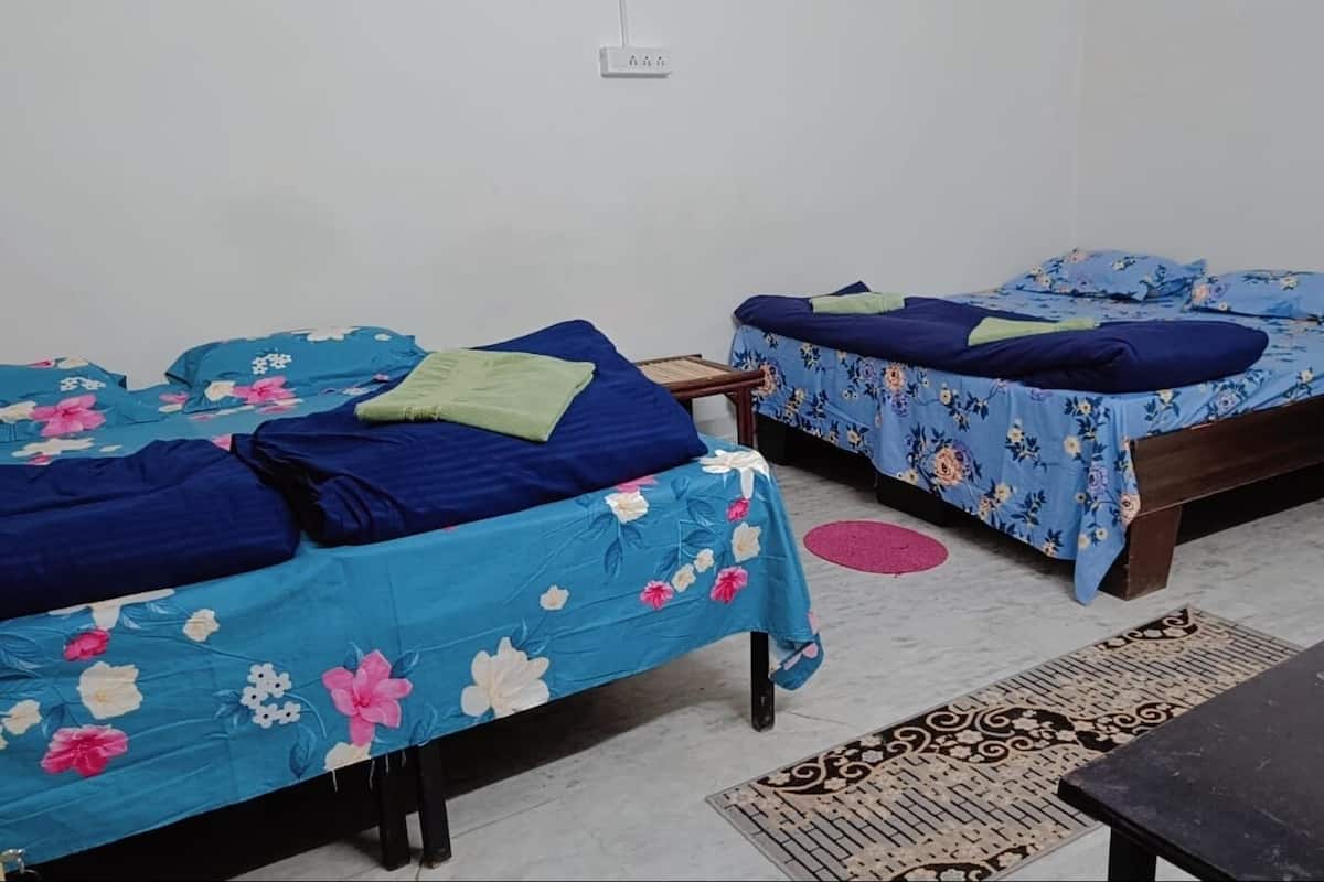 Duas camas de solteiro com colchas floridas, uma mesinha e um tapete estampado em um quarto com paredes lisas e uma tomada.