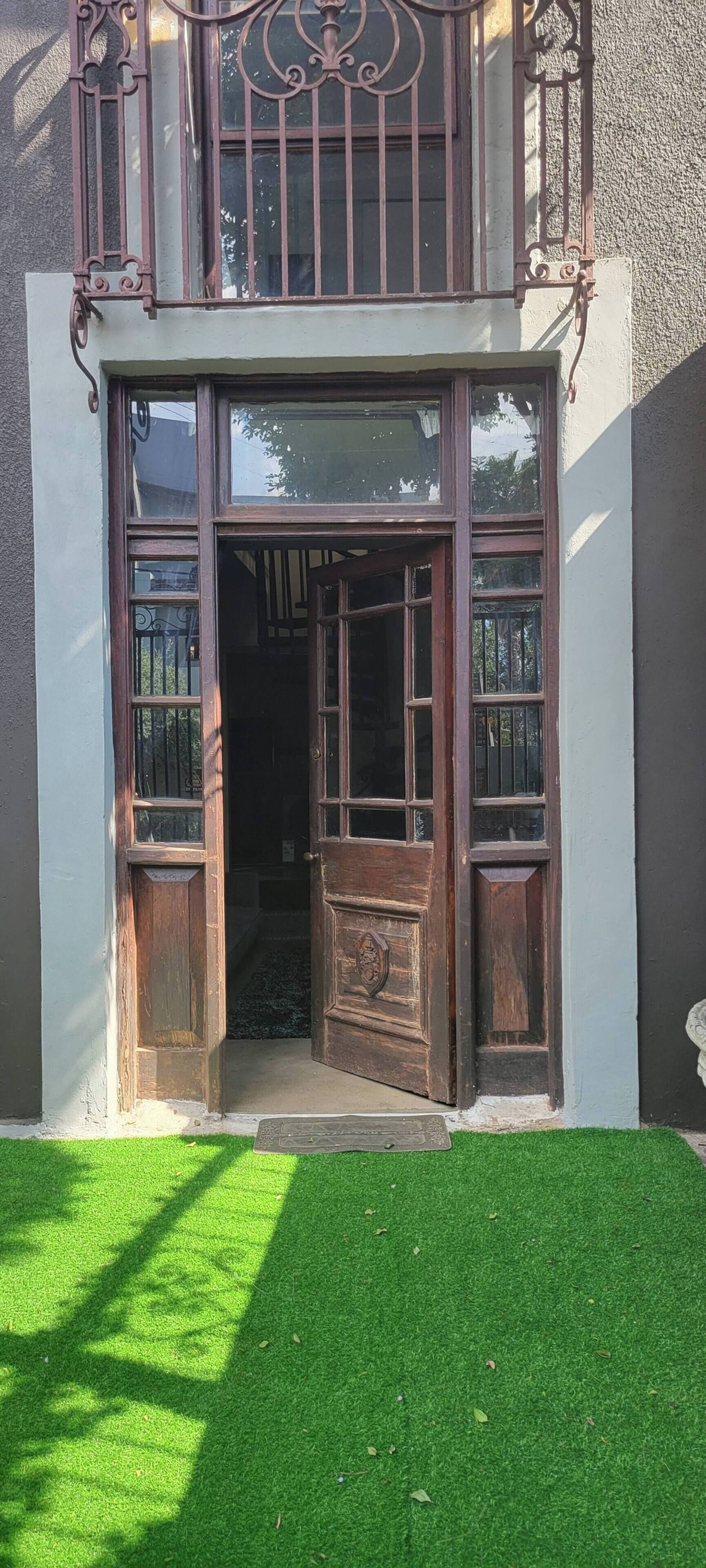 Entrada de um edifício com porta de madeira, varanda com grades de metal e um pequeno gramado na frente.