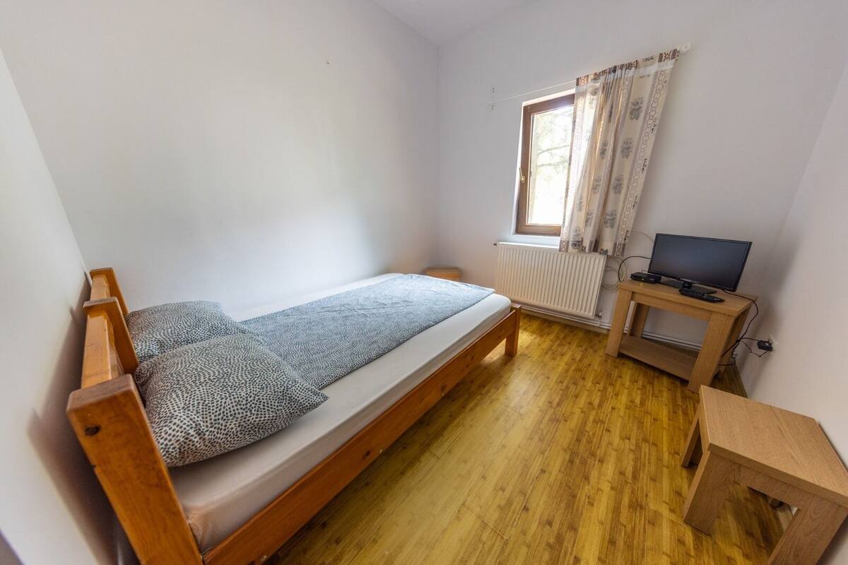 Uma cama de solteiro com estrutura de madeira, uma pequena mesa de madeira com uma televisão e uma janela com cortinas.