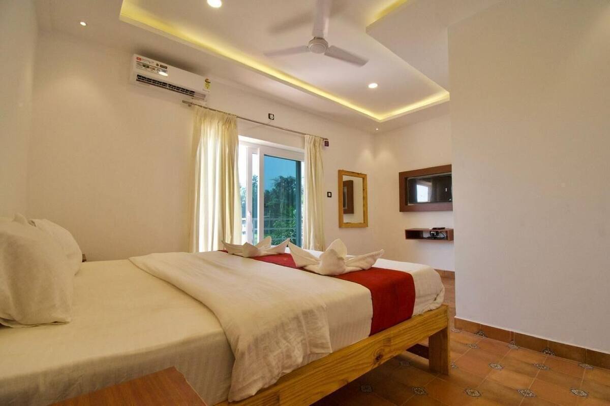 Quarto de hotel com cama, ventilador de teto, ar condicionado, janela com cortinas e televisão montada na parede.
