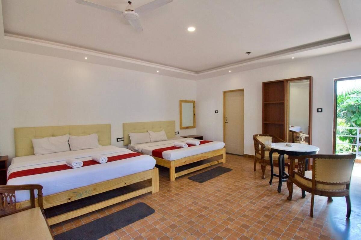 Quarto de hotel com duas camas, uma mesa de jantar, cadeiras, um ventilador de teto e uma porta que dá para uma varanda.