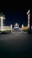 Entrada de hotel com acesso por portão, palmeiras e sinalização iluminada.