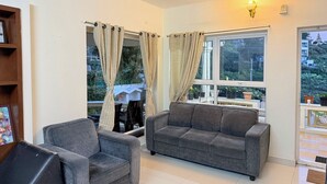 Uma sala de estar com sofá cinza, poltrona e cortinas, uma estante de madeira e uma janela com vista para o verde.