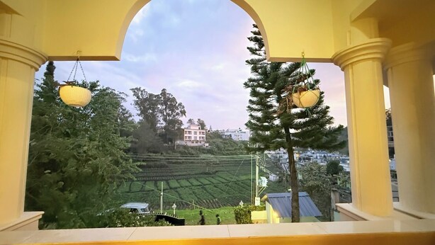 Vista através de uma janela arqueada com luminárias pendentes, um edifício ao fundo e paisagem verdejante.