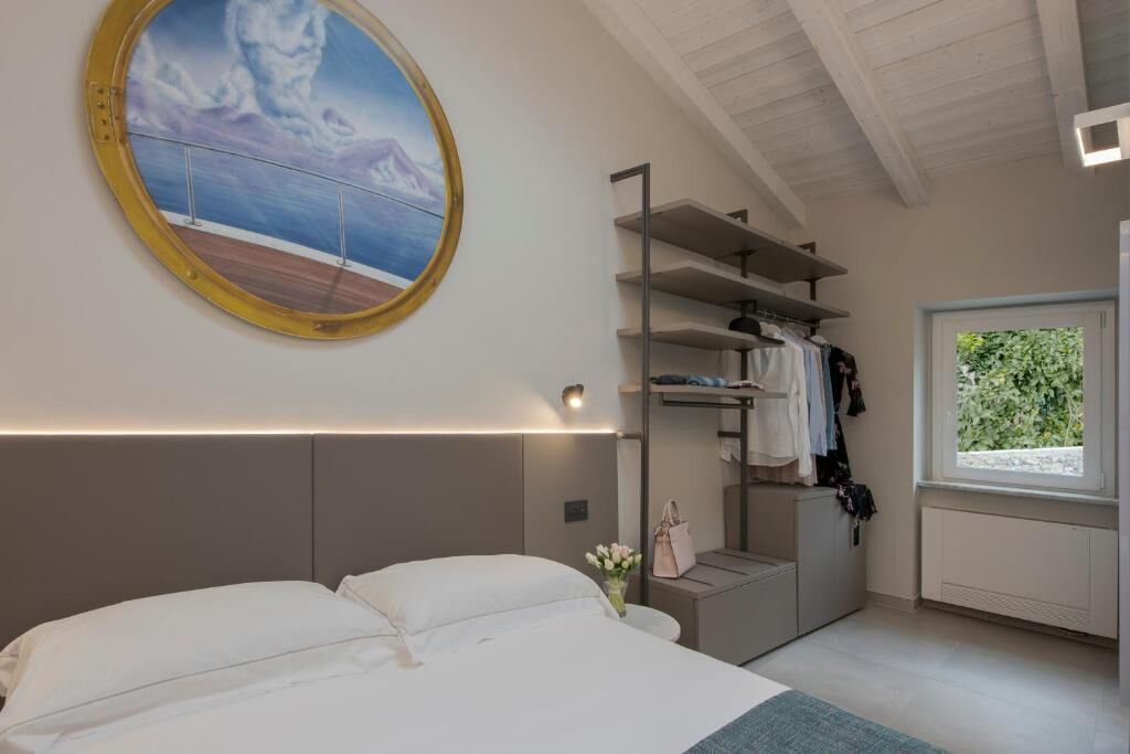 Um quarto com cama, prateleira na parede com roupas, janela com vista e um quadro.