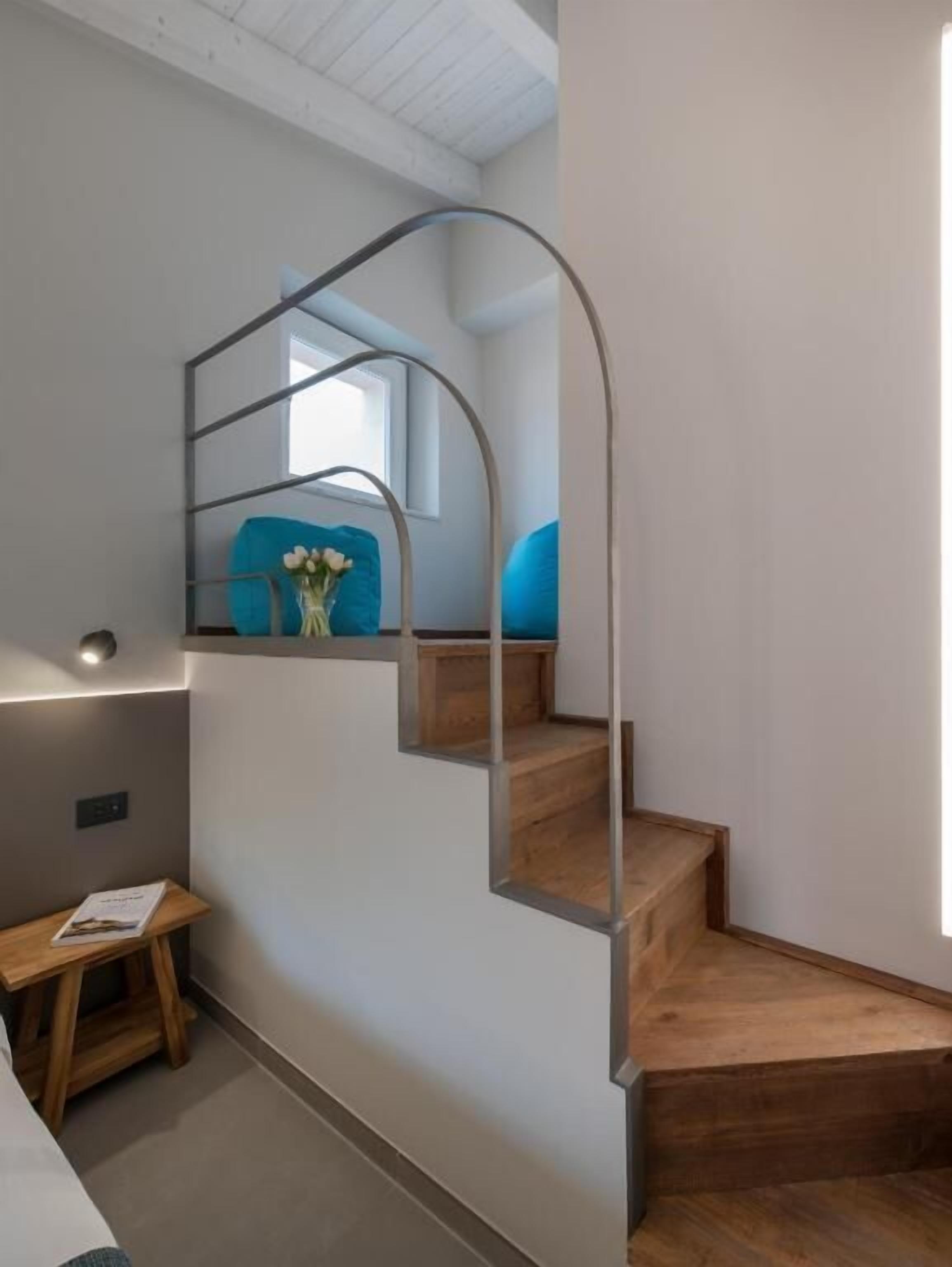 Uma escadaria moderna com corrimão de metal, uma pequena mesa com uma revista e uma almofada azul ao fundo.