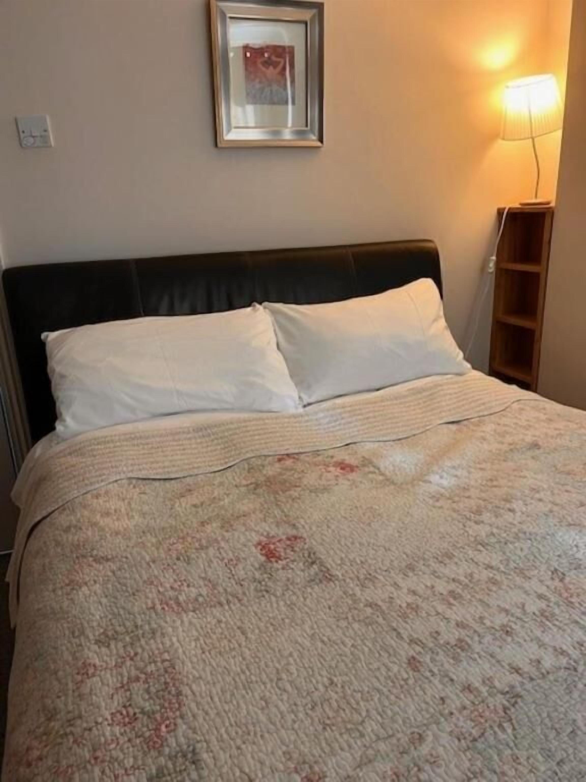 Cama com almofadas brancas e manta com padrão floral, quadro na parede e abajur.